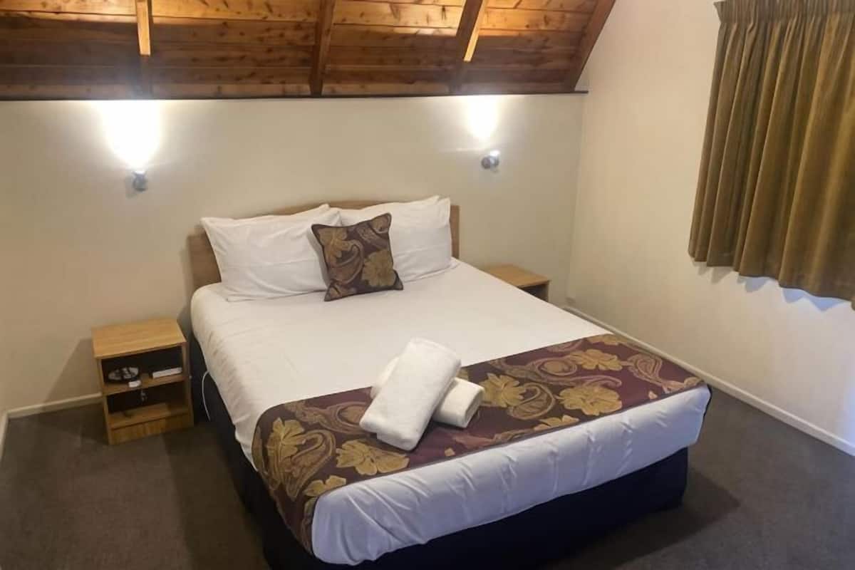 Quarto com cama, duas mesinhas de cabeceira, um criado-mudo, janela com cortinas e teto de madeira.