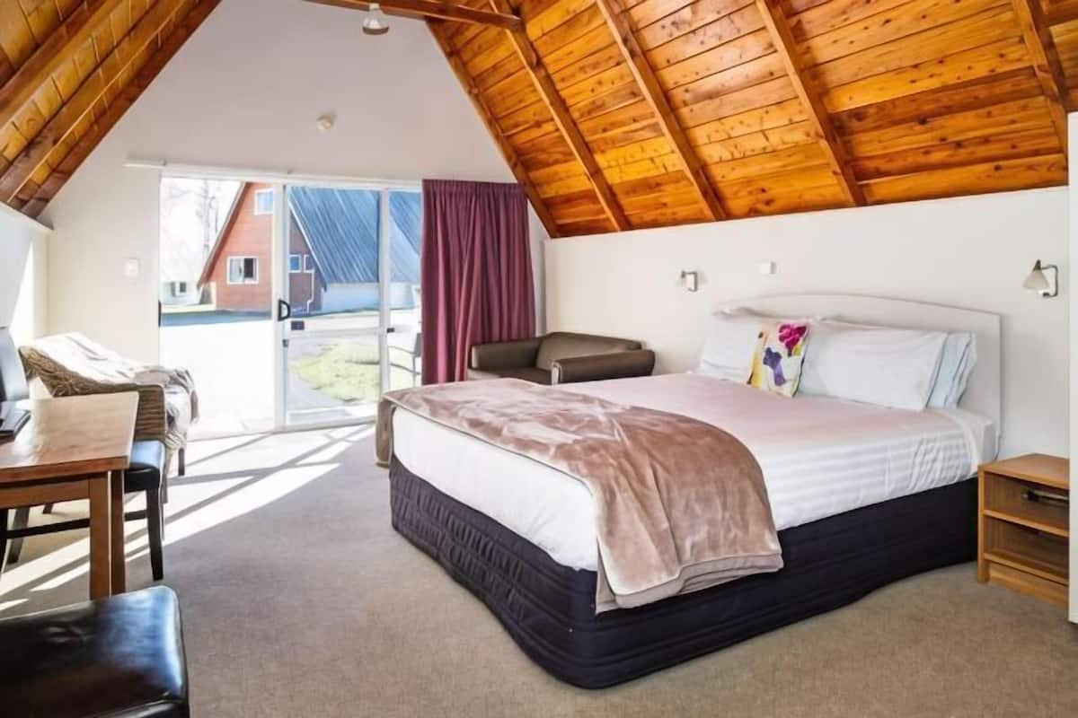 Quarto com cama grande, escrivaninha, cadeira e janela com vista para um edifício.