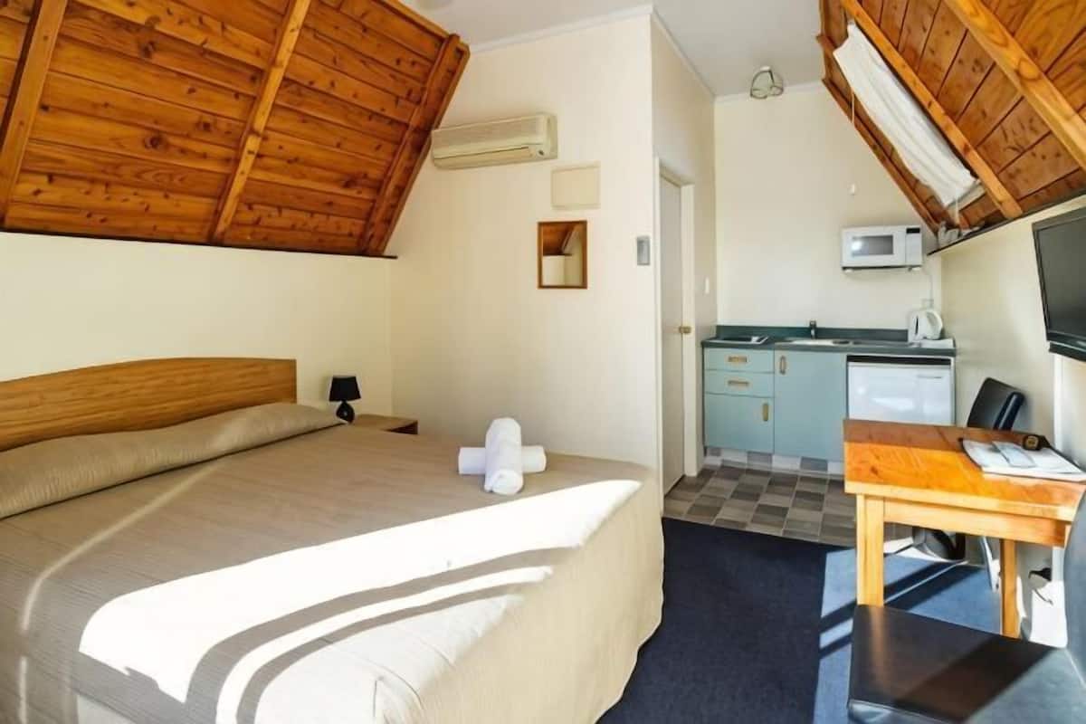 Quarto com cama, teto de madeira, cozinha compacta e uma pequena mesa.