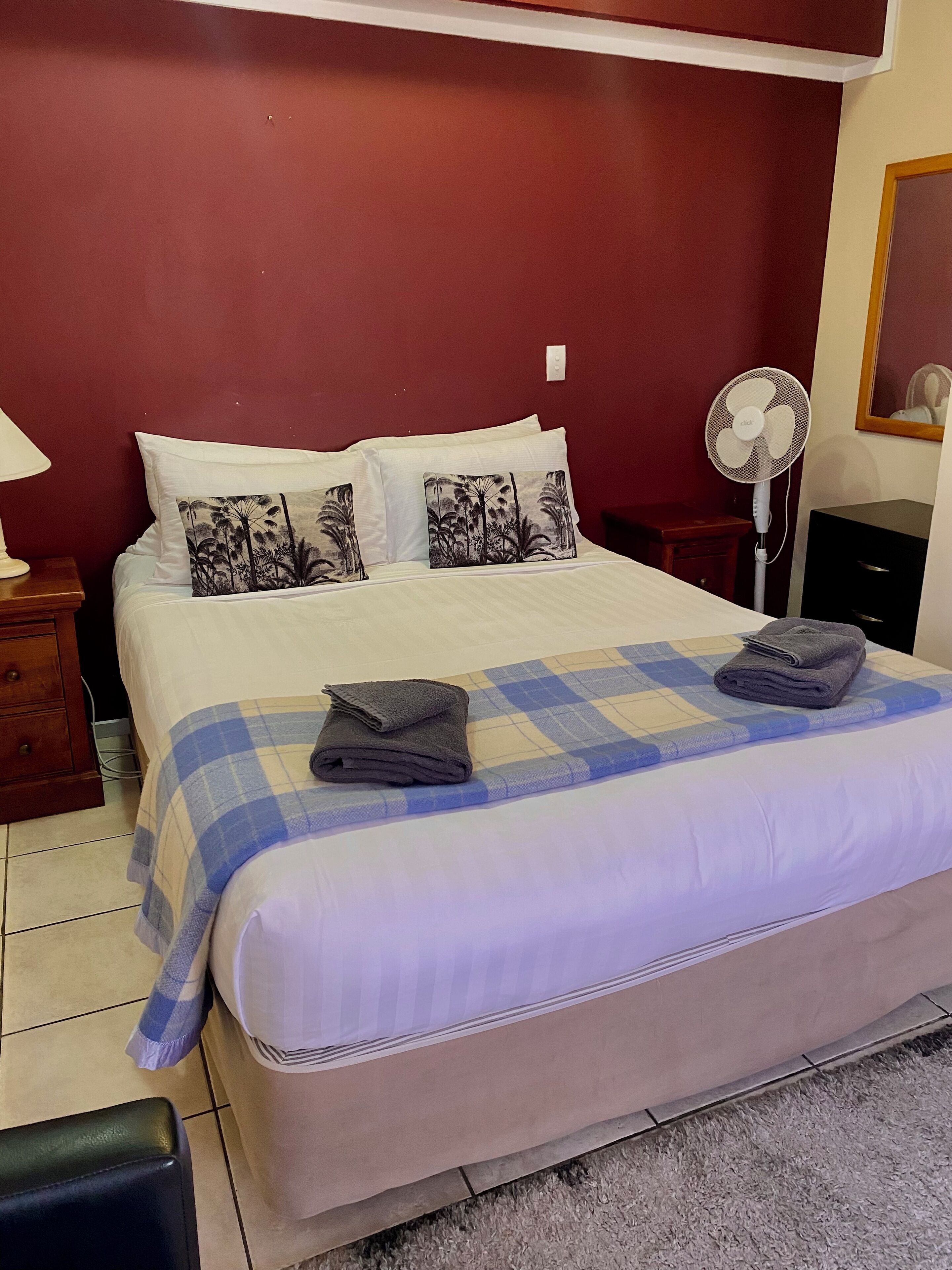 Cama bem arrumada com manta xadrez, dois travesseiros com estampas tropicais, um ventilador e uma mesa de cabeceira com luminária.