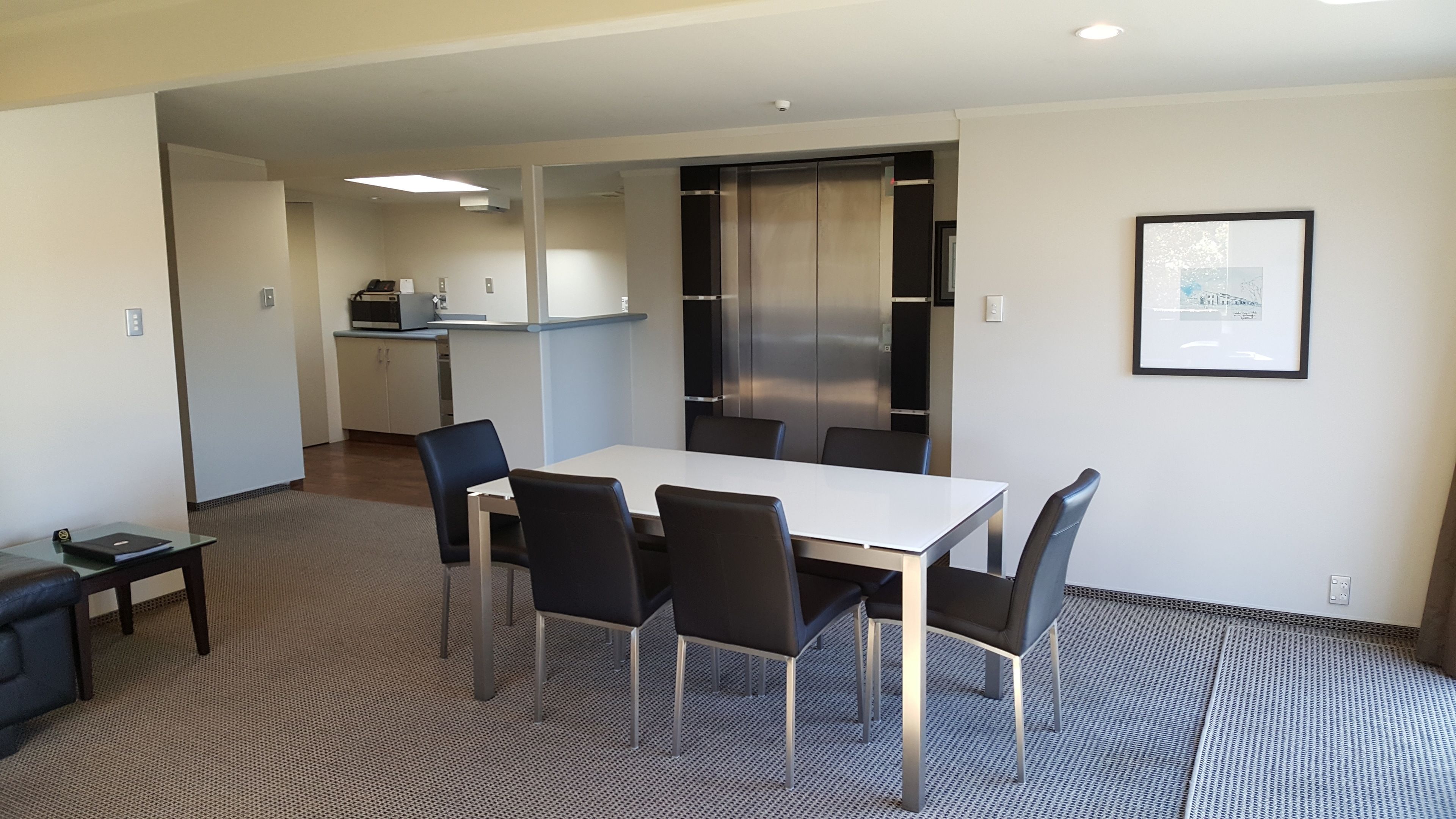 Sala de conferências com uma mesa branca e seis cadeiras, uma kitchenette ao fundo e um quadro na parede.