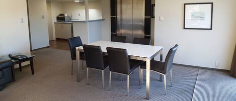 Sala de conferências com uma mesa branca e seis cadeiras, uma kitchenette ao fundo e um quadro na parede.
