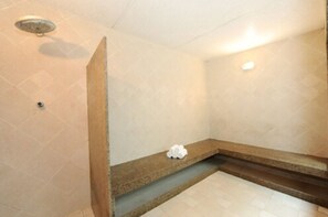 Um banheiro pequeno revestido com azulejos, com chuveiro, um banco e uma toalha no chão.
