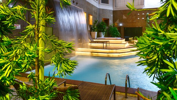 Piscina interna com efeito cascata, rodeada por espreguiçadeiras e plantas em vasos.