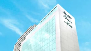 Um hotel moderno com fachada de vidro, uma placa bem visível e uma área ajardinada na base.