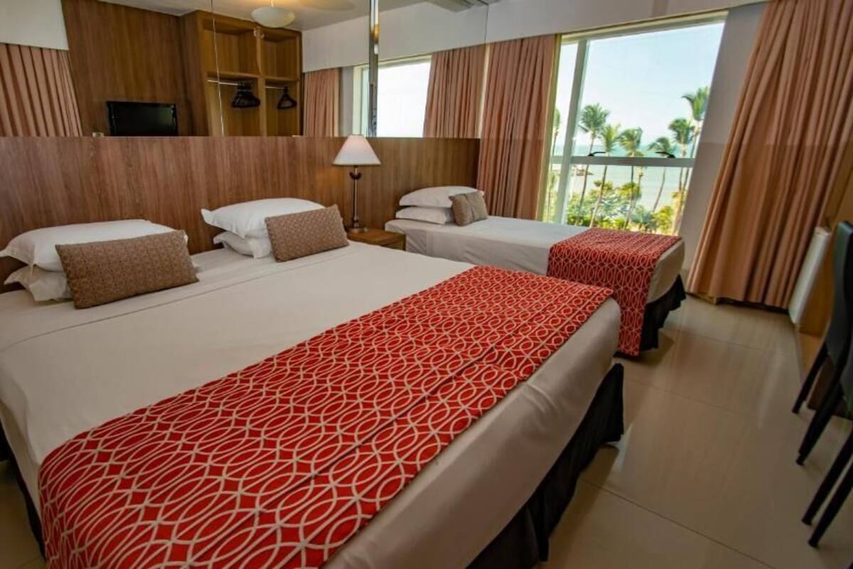Quarto de hotel com duas camas, televisão, varanda com cortinas e parede de madeira.
