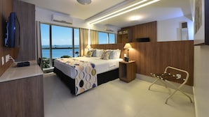Quarto de hotel moderno com uma cama grande, luminárias laterais, uma TV e vista para o mar.