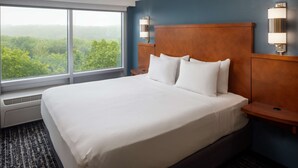 Quarto de hotel com uma cama grande, duas mesas de cabeceira, um criado-mudo, uma janela com vista para o verde e luminárias fixadas na parede.