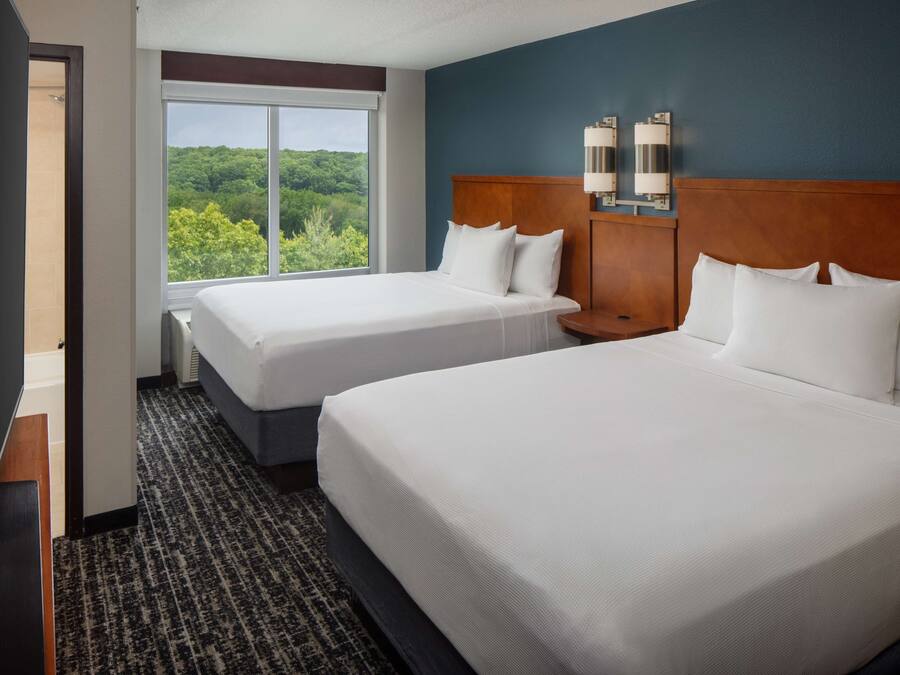 Quarto de hotel com duas camas, uma janela ampla com vista para área verde e cabeceira de madeira.