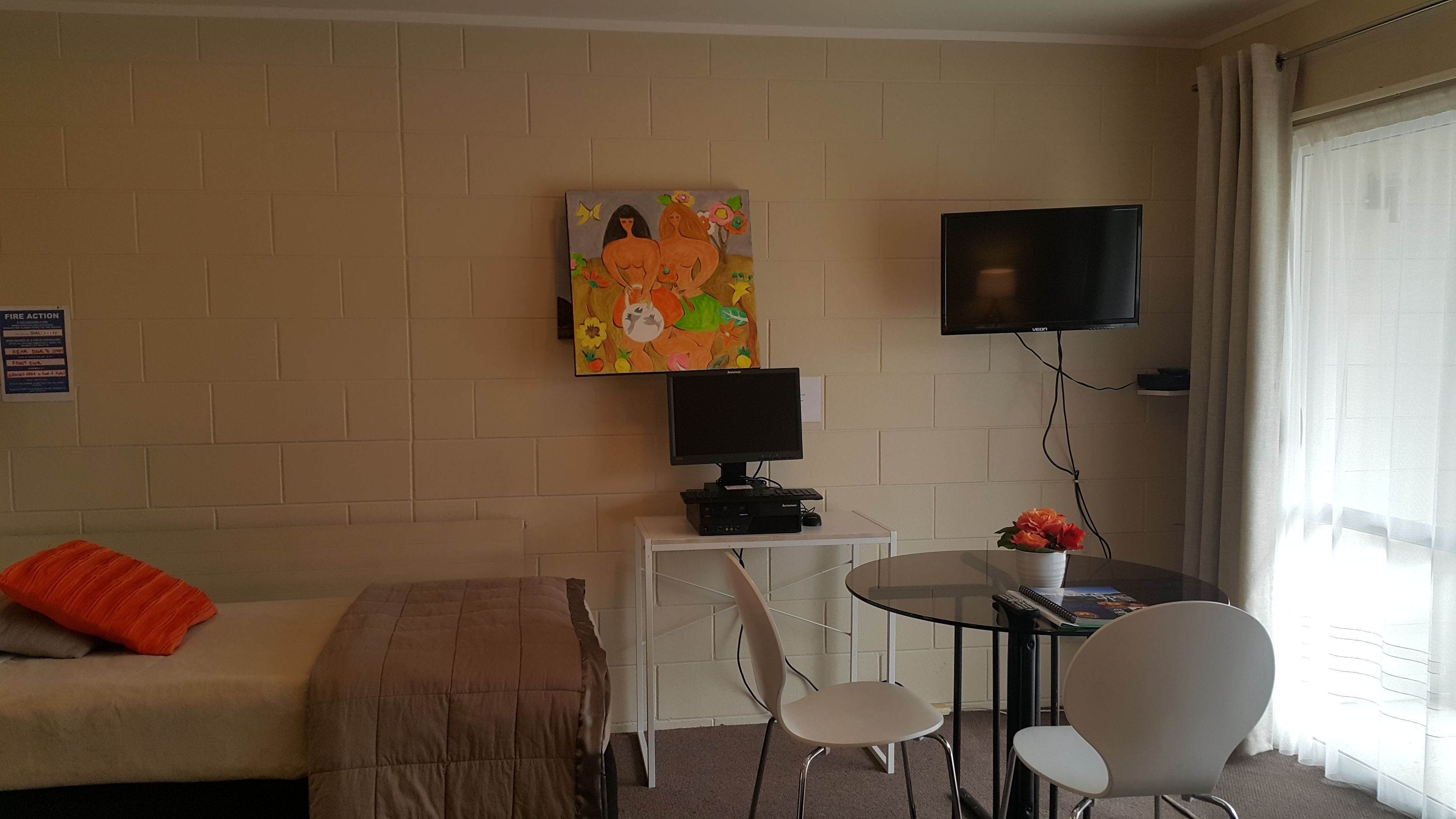 Quarto de hotel com uma cama, uma mesa pequena com uma televisão, um quadro na parede e uma janela com cortinas.
