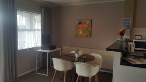 Quarto compacto com uma mesa de jantar, duas cadeiras, pequena cozinha com chaleira e um quadro na parede.