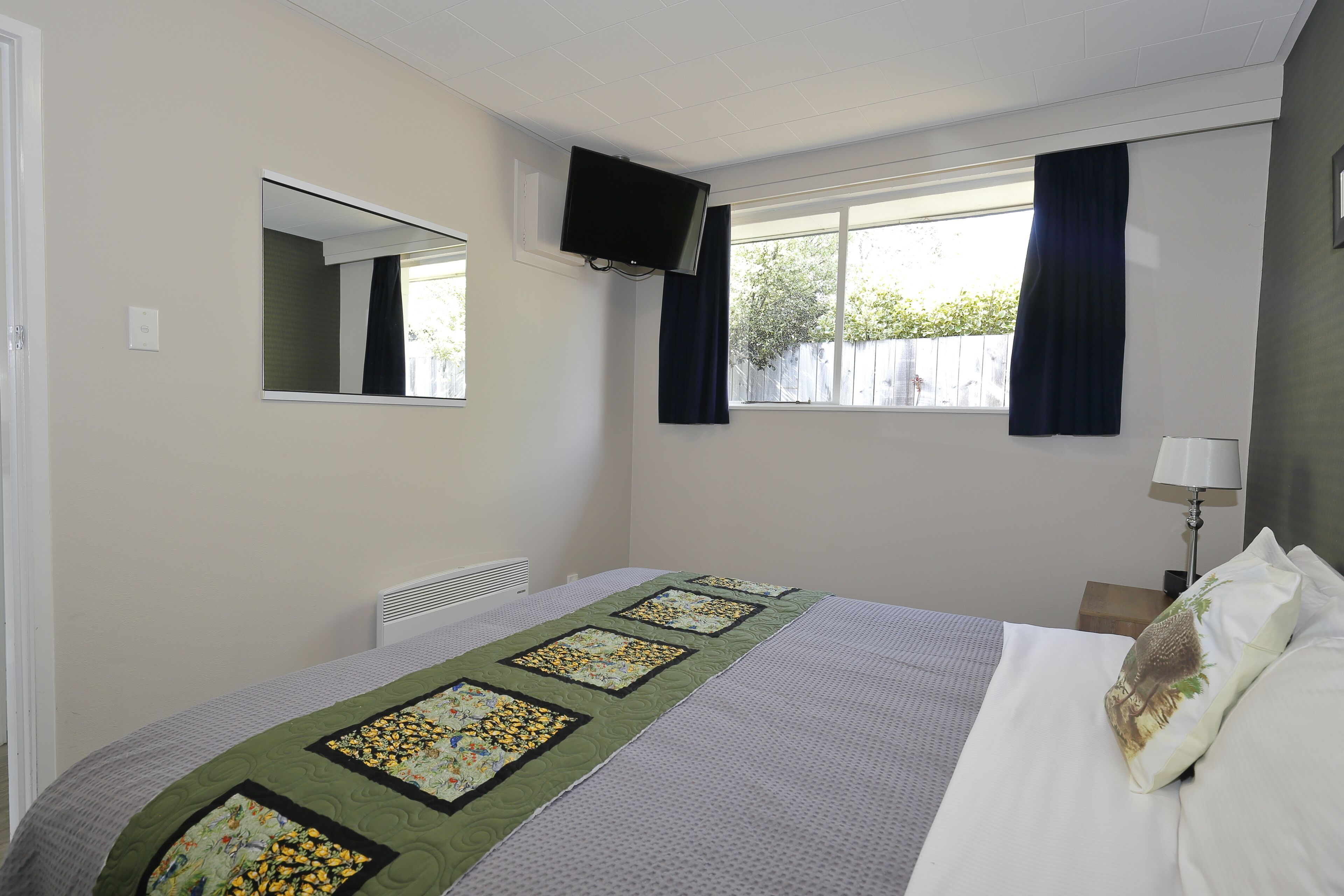 Quarto de hotel com cama, televisão na parede, janela com cortinas e mesa de cabeceira com abajur.