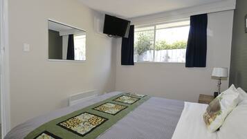 Quarto de hotel com cama, televisão na parede, janela com cortinas e mesa de cabeceira com abajur.