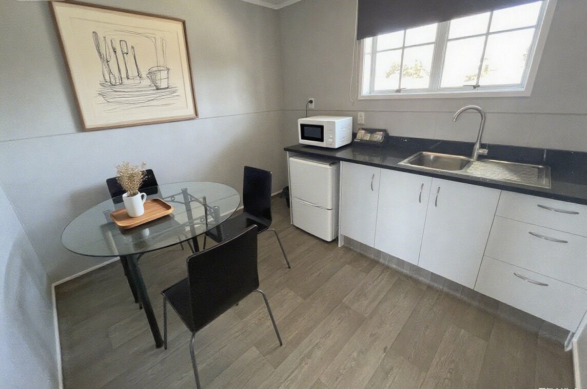 Uma cozinha compacta com micro-ondas, pia e um pequeno frigorífico. Há uma mesa de jantar com cadeiras e uma janela que proporciona luz natural.