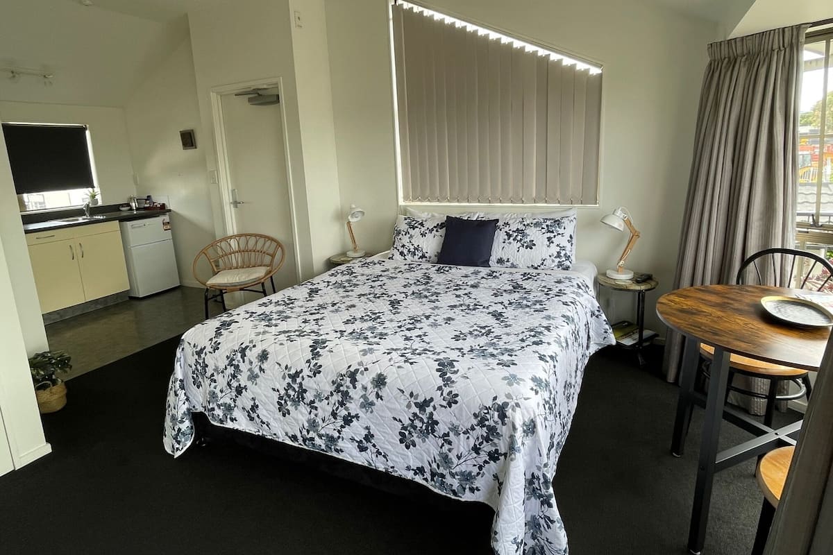 Quarto de hotel com cama, pequena mesa, cadeira, janela com cortinas, porta e cozinha compacta.