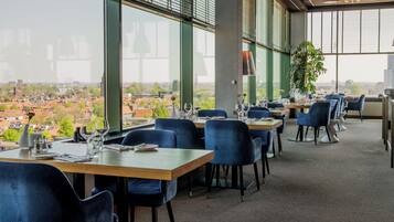 Um restaurante com grandes janelas que oferecem vista para a cidade, mesas montadas com taças de vinho e pratos, e cadeiras estofadas azuis.