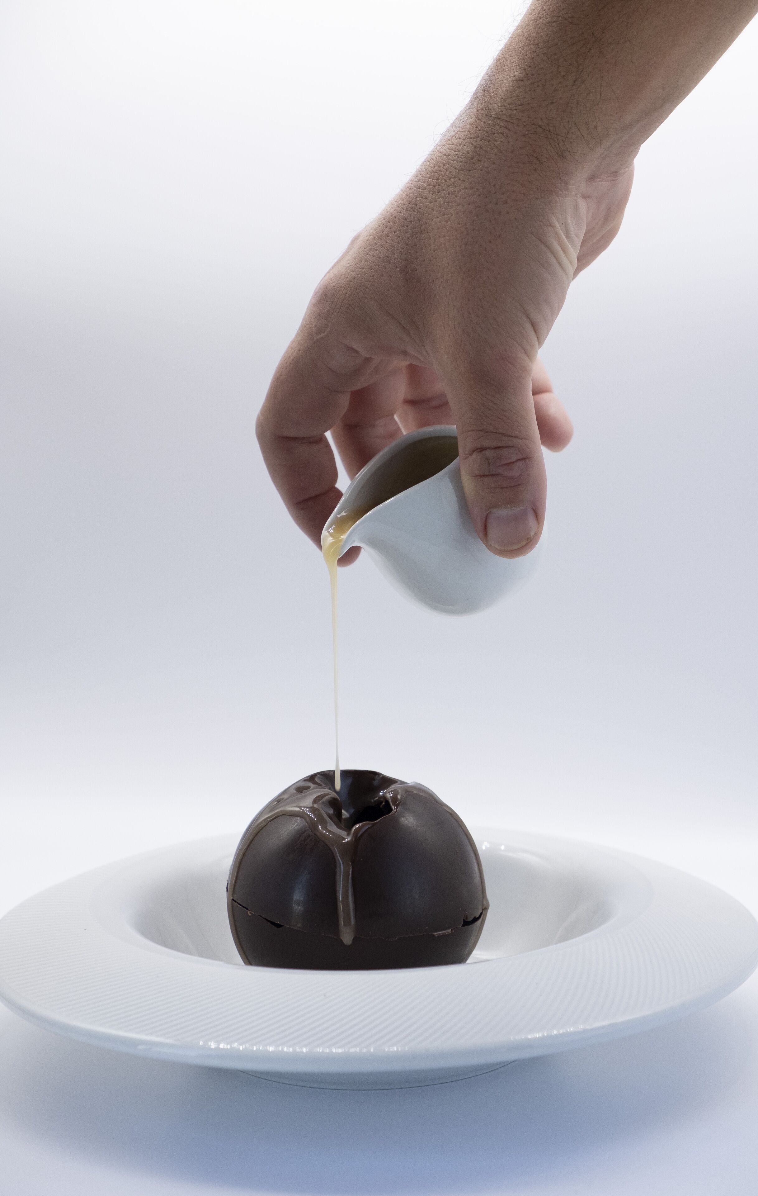 Uma mão despejando líquido de uma xícara sobre uma esfera coberta de chocolate em um prato.