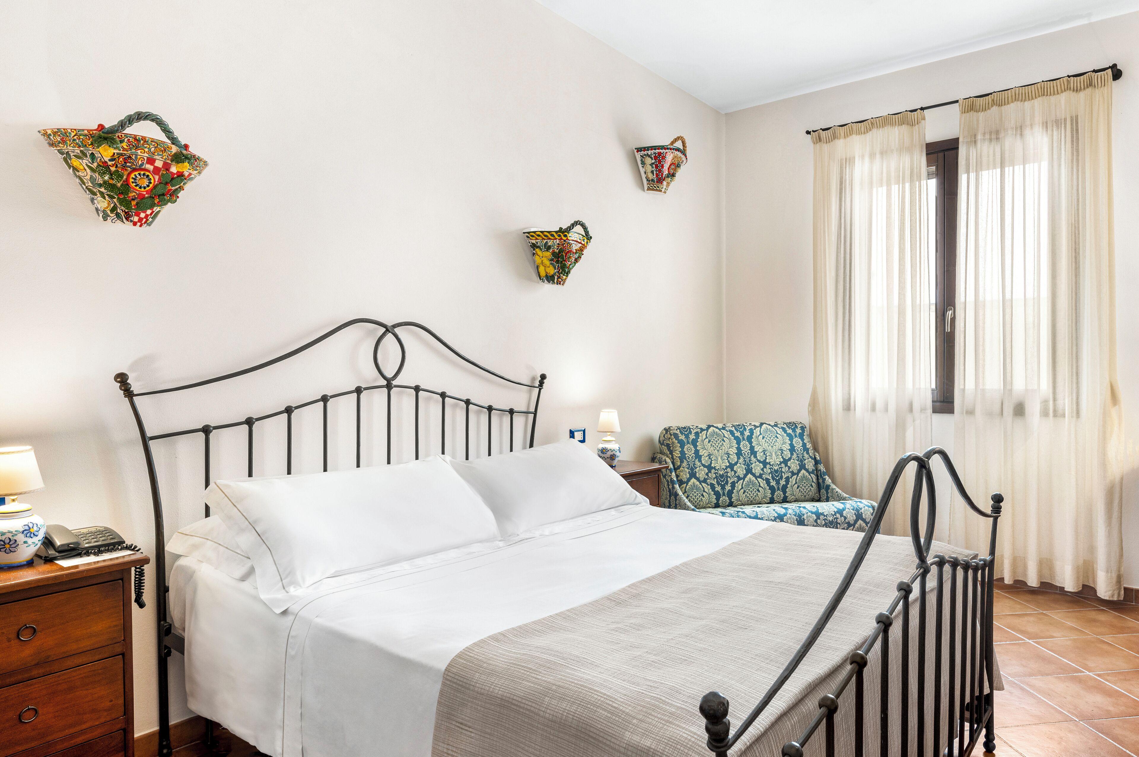 Uma cama bem arrumada com lençóis brancos, uma mesa de cabeceira com abajur e uma cadeira com almofada estampada.