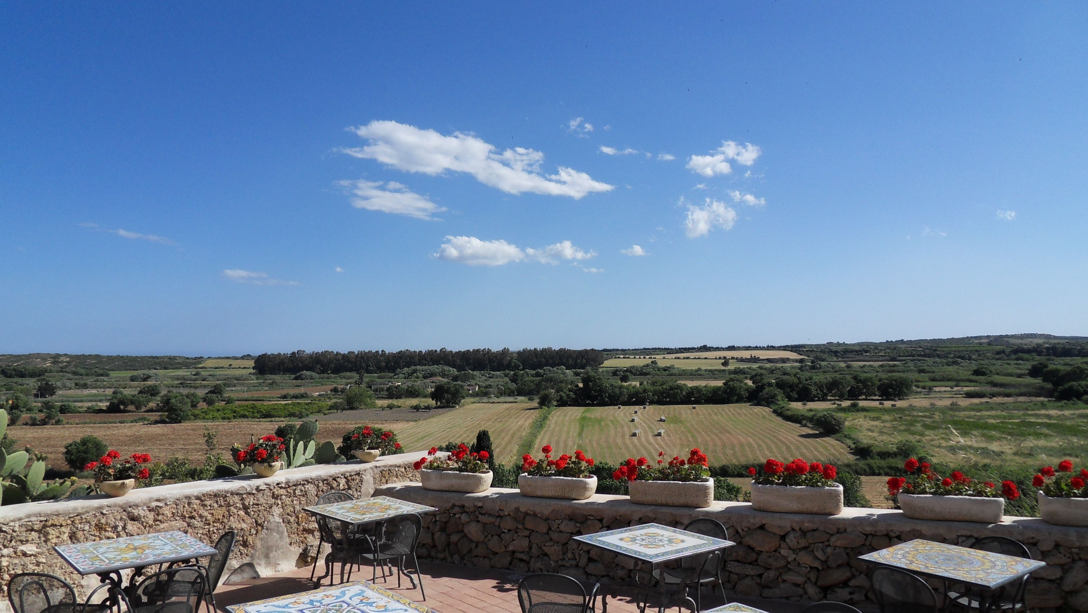 Um terraço com paredes de pedra, mesas e cadeiras ao ar livre, com vista para uma paisagem rural com campos e árvores sob um céu claro.