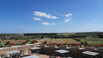 Um terraço com paredes de pedra, mesas e cadeiras ao ar livre, com vista para uma paisagem rural com campos e árvores sob um céu claro.