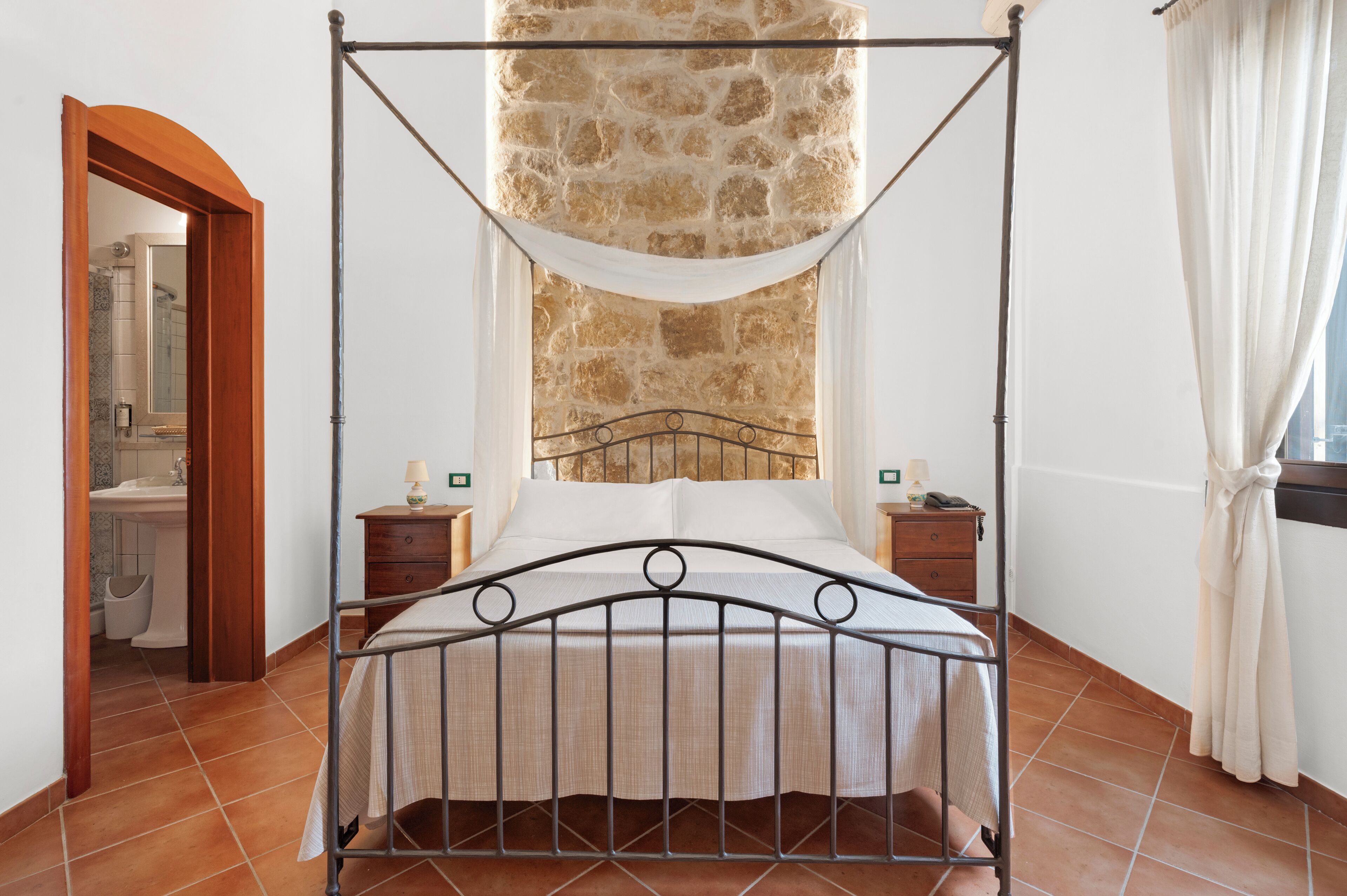 Uma cama com dossel, parede de pedra ao fundo, porta de madeira para um banheiro e janela com cortinas.