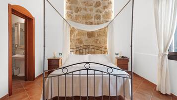 Uma cama com dossel, parede de pedra ao fundo, porta de madeira para um banheiro e janela com cortinas.