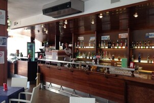 Uma área de bar com balcão de madeira, prateleiras com garrafas e uma pessoa atrás do balcão.
