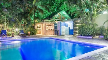 Uma área de piscina com piscina azul cristalina, cercada por vegetação exuberante e palmeiras. Há espreguiçadeiras e uma pequena estrutura com porta de vidro, provavelmente um bar ou lounge à beira da piscina.