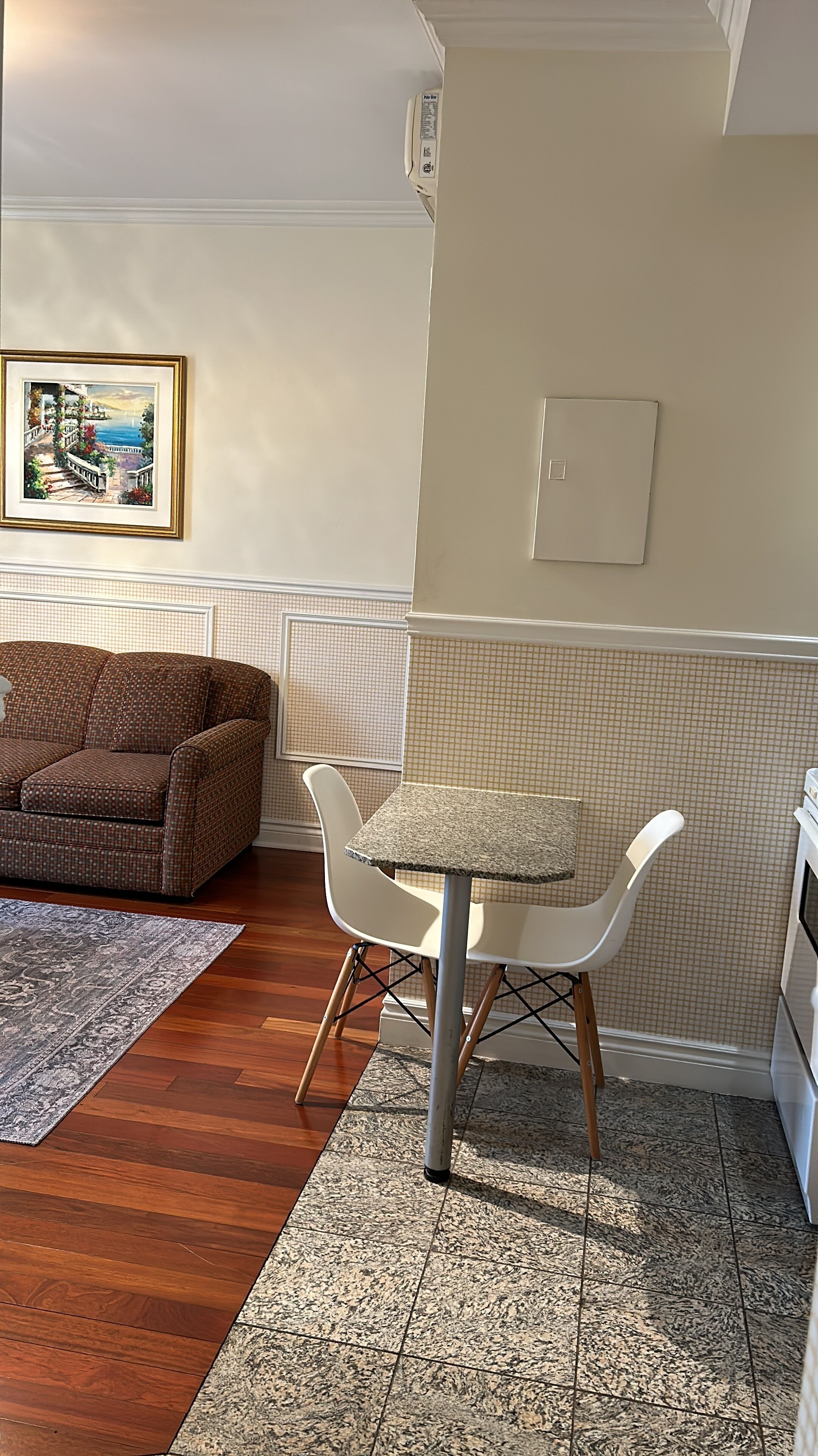 Uma sala de estar com um sofá de padrão marrom, uma mesinha redonda com uma cadeira branca, um quadro na parede e piso de azulejos.