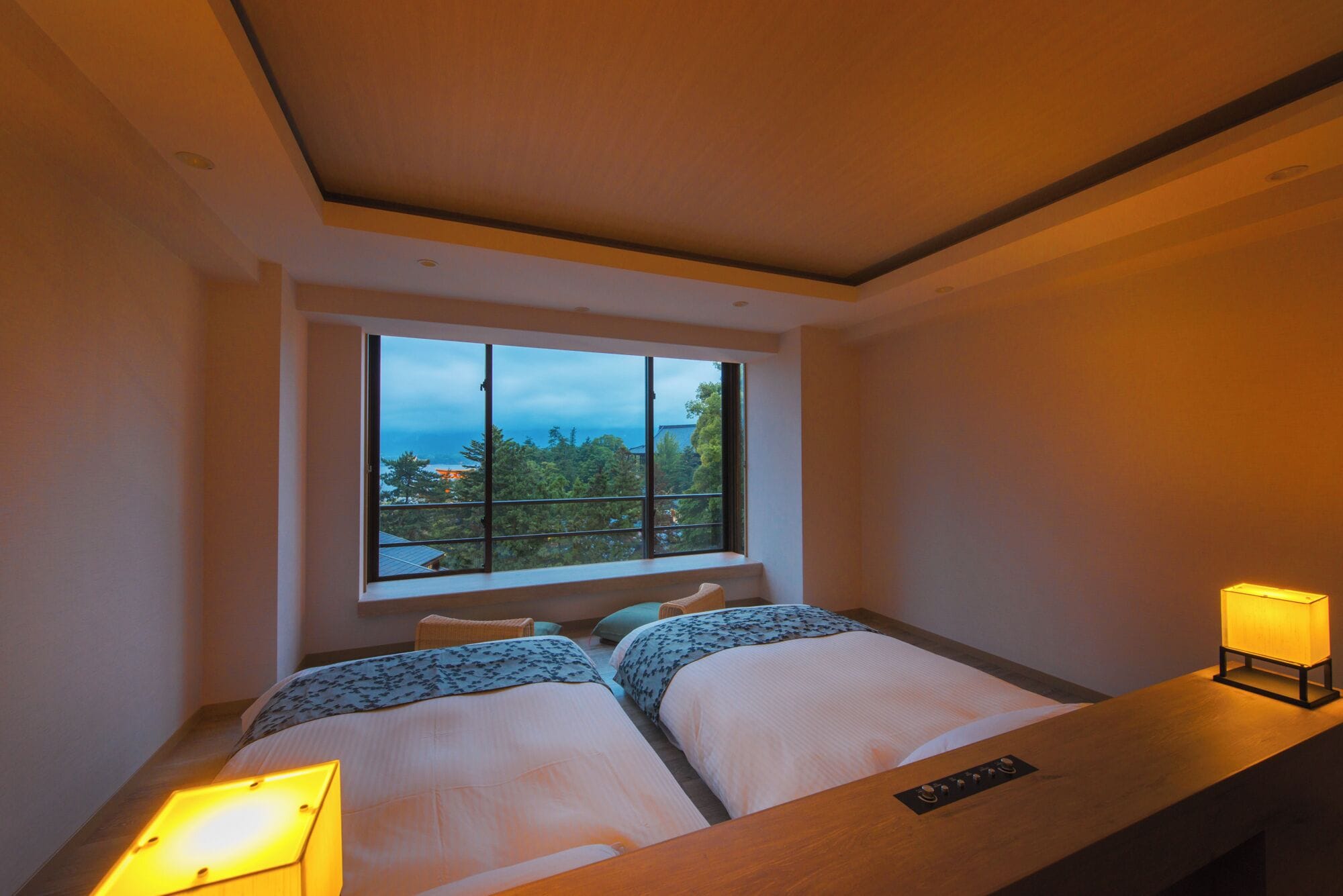 Quarto de hotel com duas camas, janela com vista para árvores e edifícios, e mesa de cabeceira com abajur.