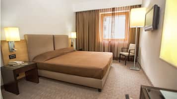 Quarto de hotel com uma cama grande, duas luminárias laterais, uma escrivaninha com telefone, uma cadeira e uma TV fixada na parede.