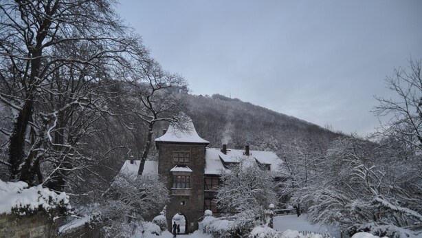 Um edifício coberto de neve com fachada de pedra e uma torre central, cercado por árvores carregadas de neve e uma colina ao fundo.