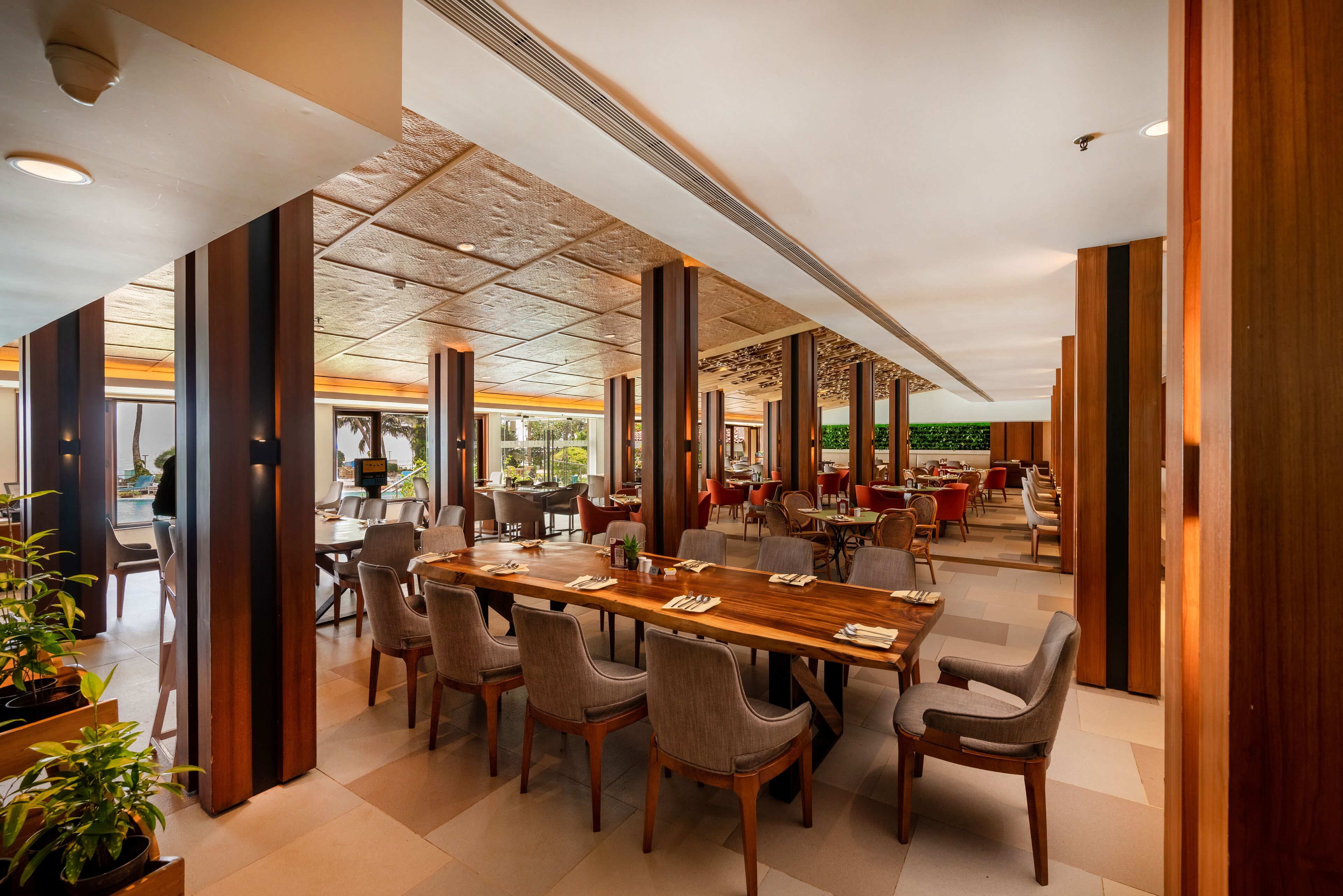 Uma sala de jantar ampla, com uma longa mesa de madeira, várias cadeiras e vista para a área externa com mesas.