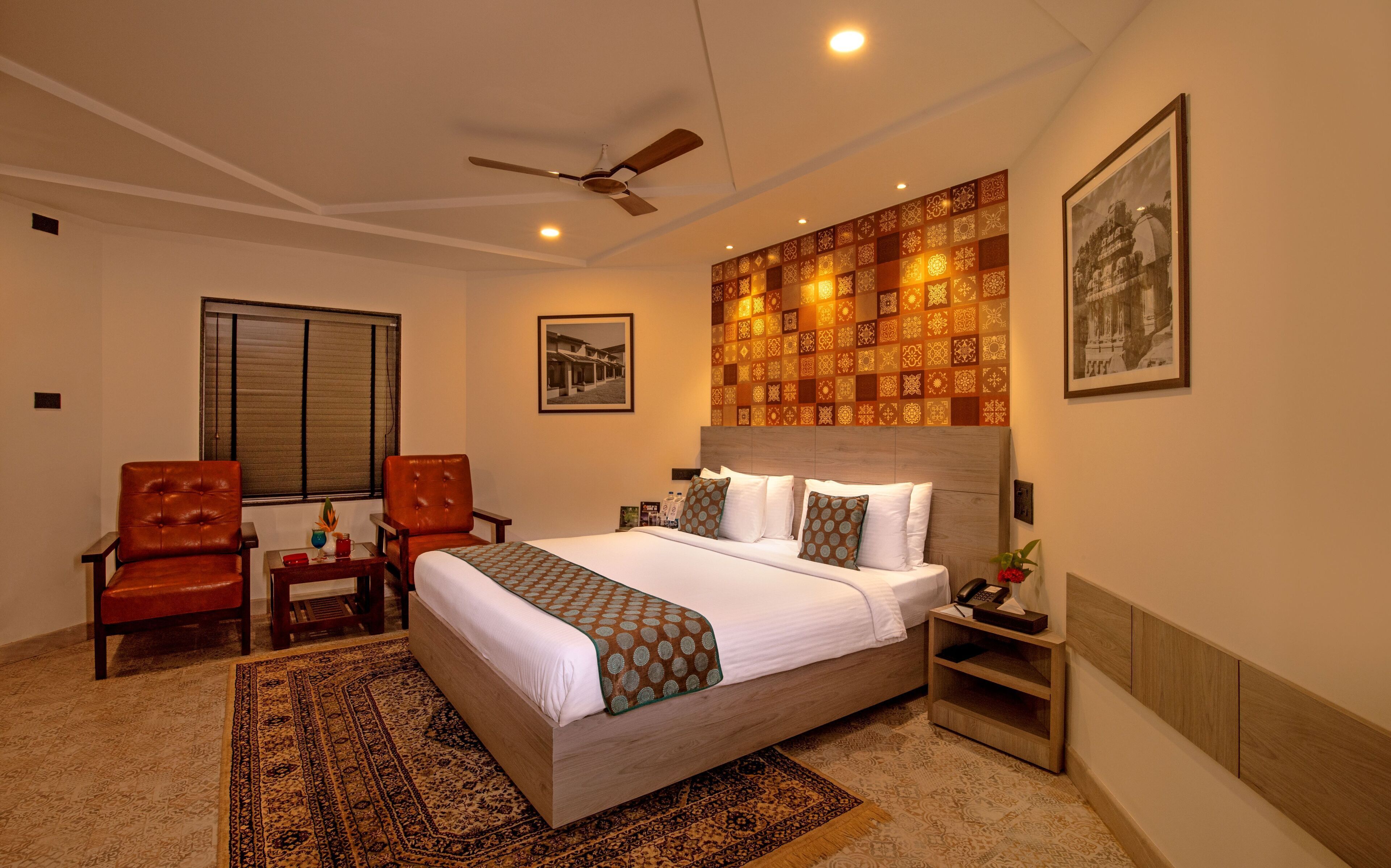 Quarto com cama, duas cadeiras, ventilador de teto, mesa de cabeceira e parede com azulejos estampados.