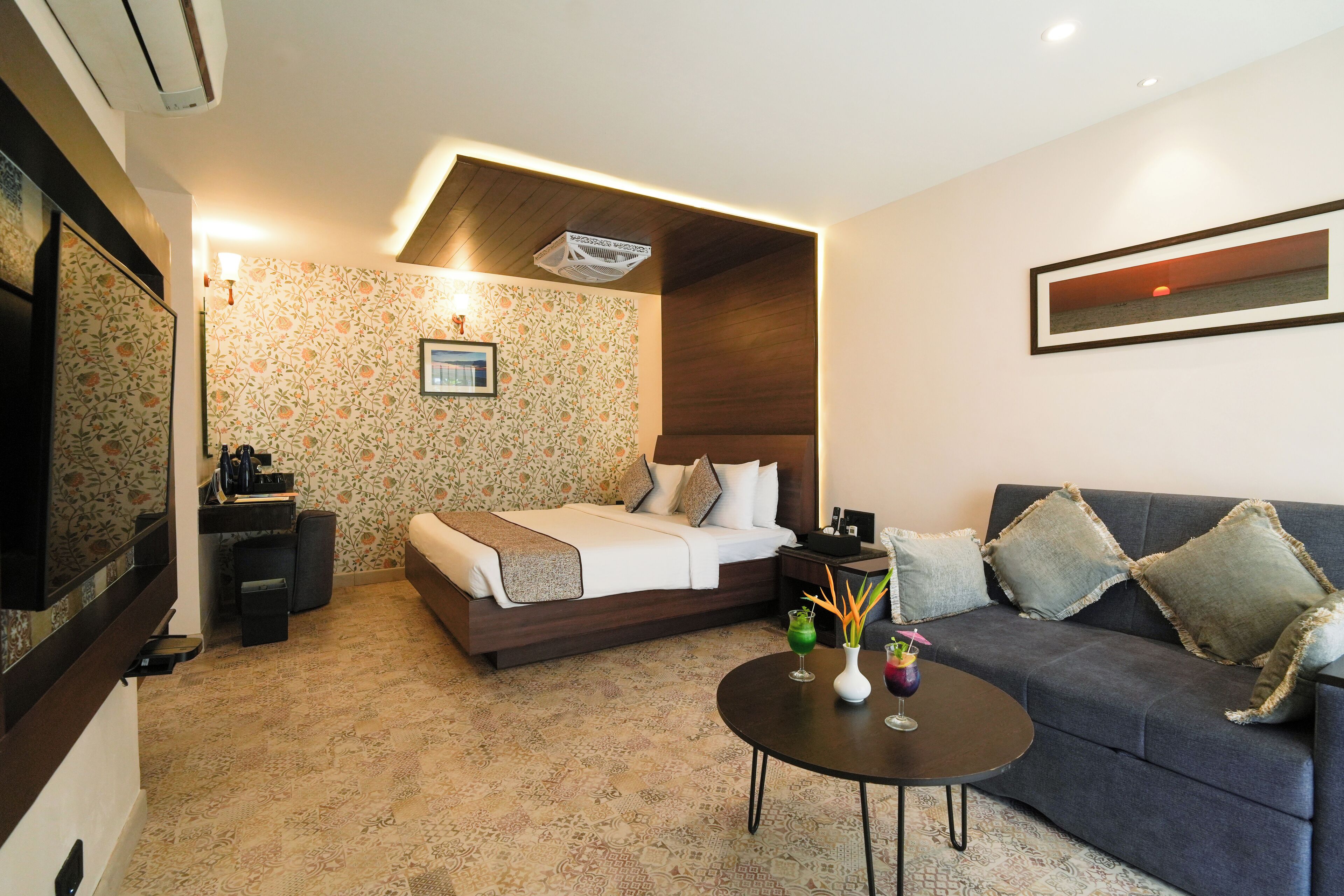 Quarto de hotel com cama, sofá, mesa de centro com bebidas, televisão e carpete estampado.