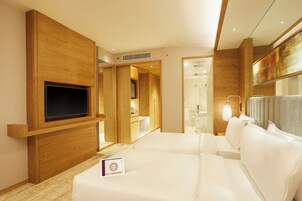 Quarto de hotel moderno com uma cama grande, televisão de tela plana fixada em um painel de madeira, closet espaçoso e um banheiro visível através de uma porta aberta.