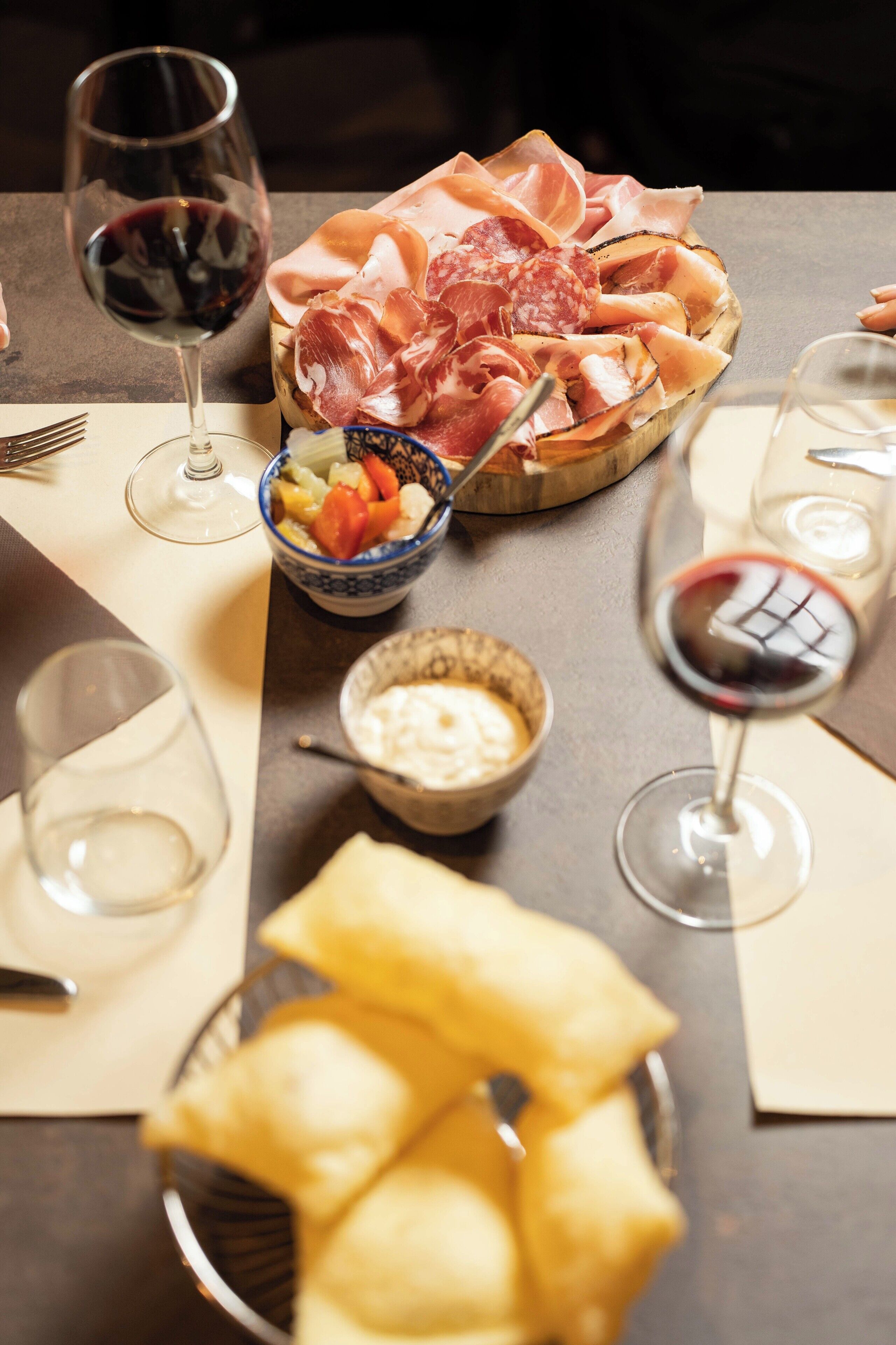 Uma mesa posta com uma tábua de frios, potes de patês e copos de vinho tinto.