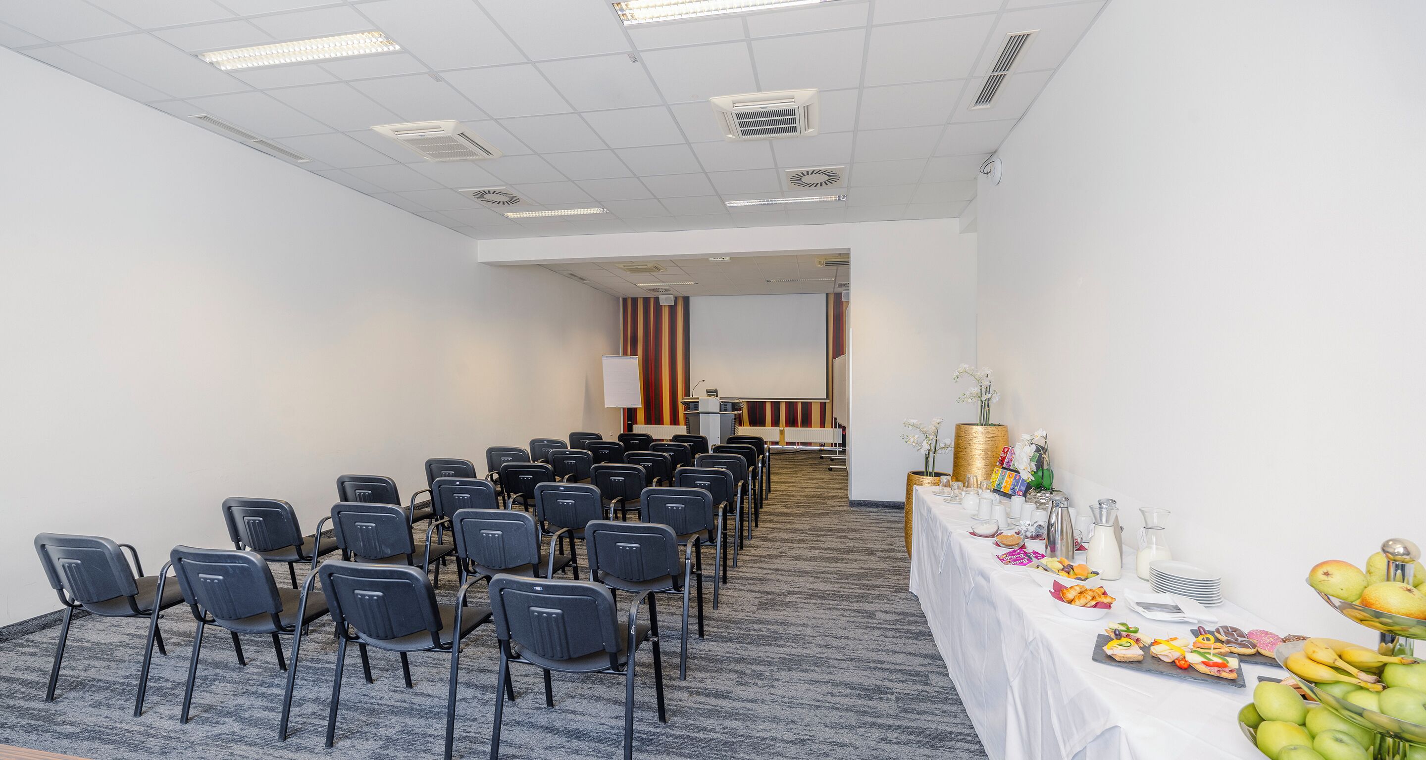 Uma sala de conferências com fileiras de cadeiras, uma mesa branca com comida e bebidas, e um púlpito na frente.