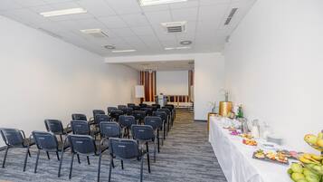 Uma sala de conferências com fileiras de cadeiras, uma mesa branca com comida e bebidas, e um púlpito na frente.