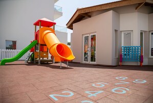 Um parquinho com um escorrega e uma estrutura de escalada, uma casinha azul e laranja, e um edifício com fachada branca e porta com painéis de vidro.