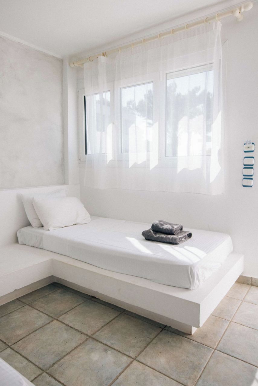 Quarto minimalista com uma cama branca, uma mesa pequena e uma janela com cortinas finas.