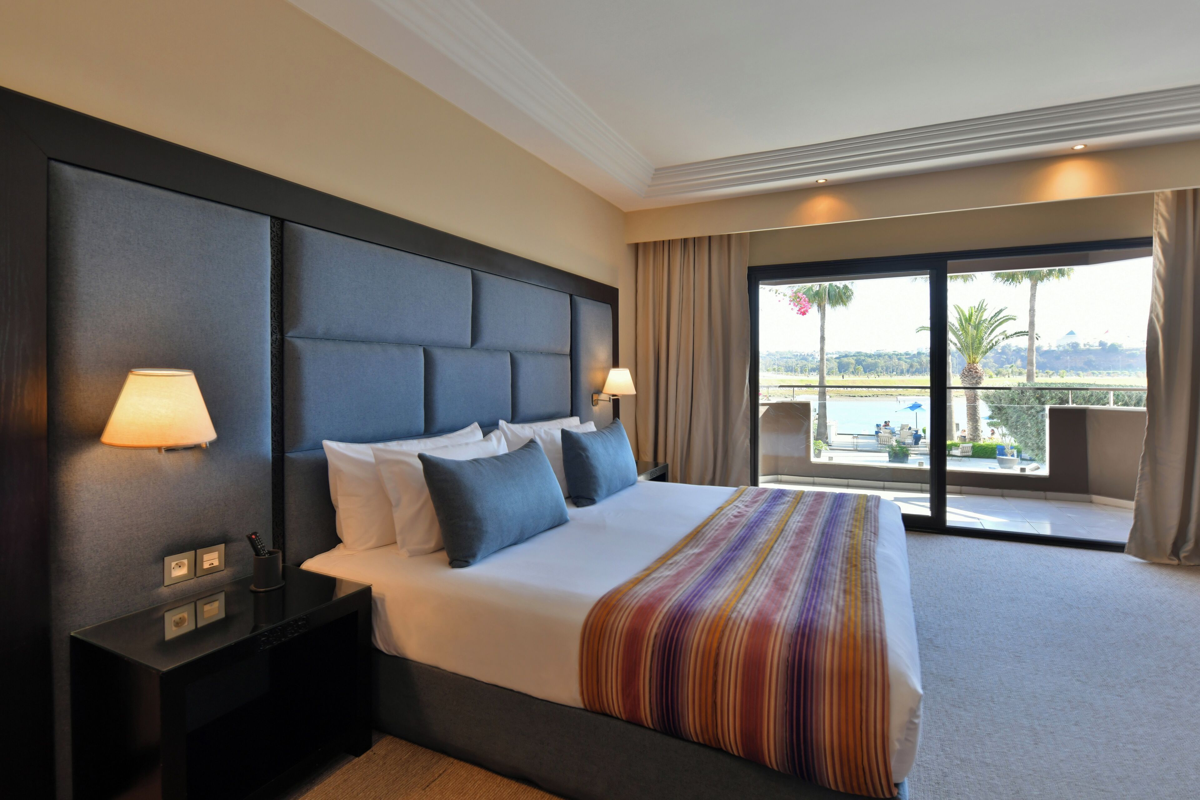 Quarto de hotel com uma cama grande, duas mesas de cabeceira, vista para o exterior e uma varanda com mesa e cadeiras.
