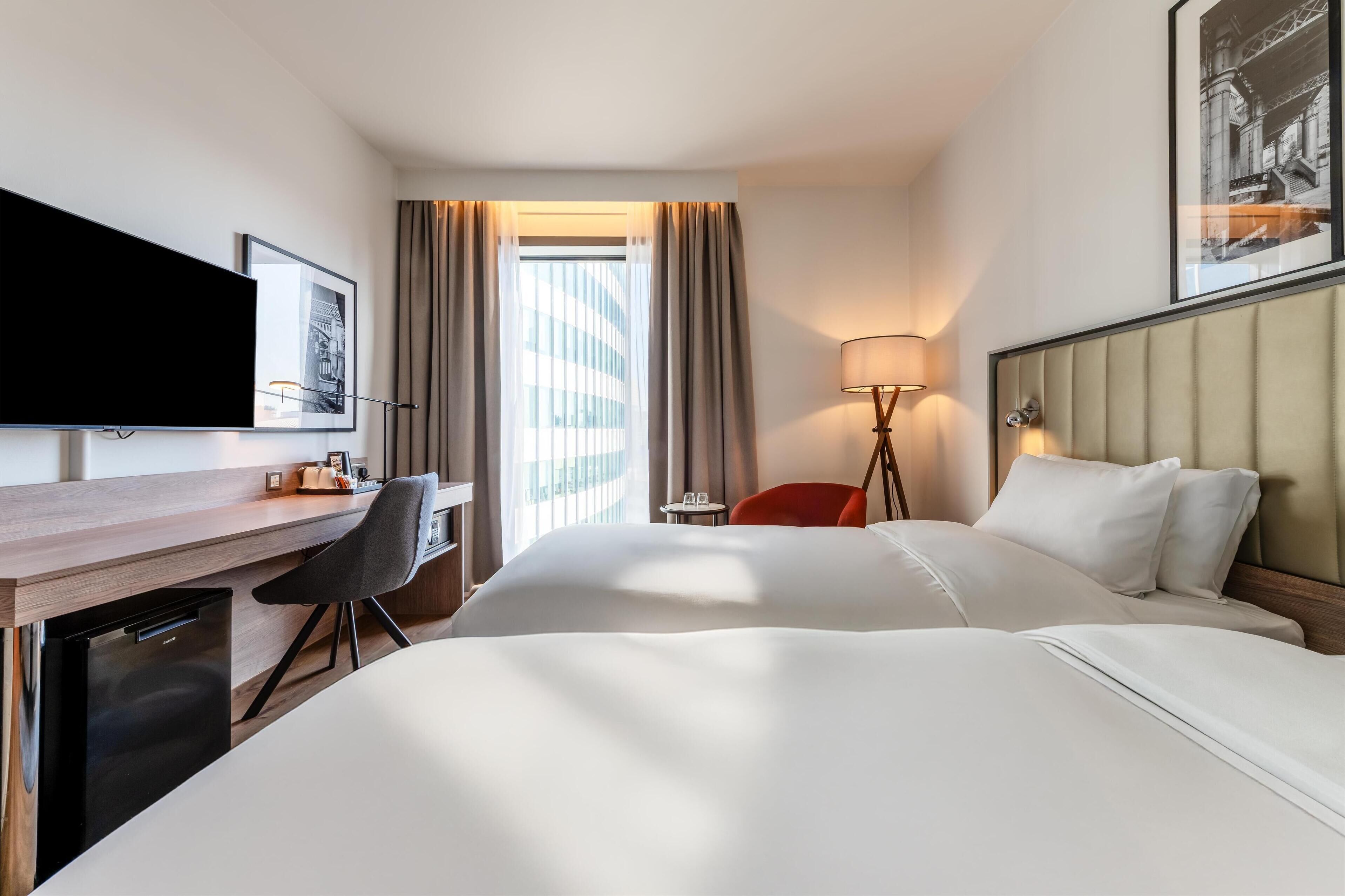 Quarto de hotel com uma cama grande, uma escrivaninha com cadeira, televisão, mesa de cabeceira, abajur e janela com cortinas.