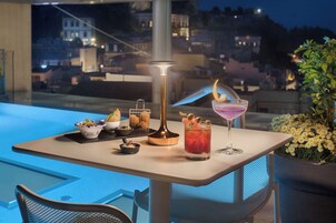 Mesa com bebida, luminária e tigela de frutas numa varanda com vista para a cidade à noite.