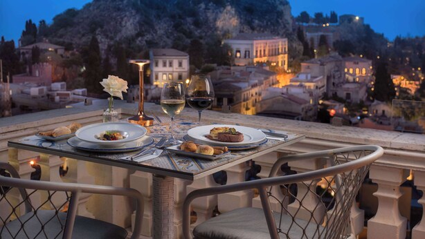 Mesa posta para jantar, com pratos de comida, copos de vinho e uma vela, com vista para a cidade ao anoitecer.