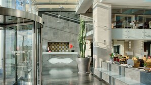 Um lobby moderno com um balcão de recepção, uma grande planta em vaso e um elevador de vidro.