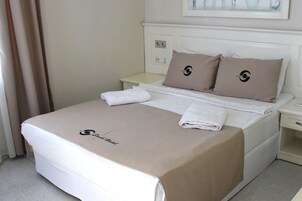 Cama bem arrumada com colcha bege e duas almofadas, mesa de cabeceira branca com abajur, e um quadro na parede.