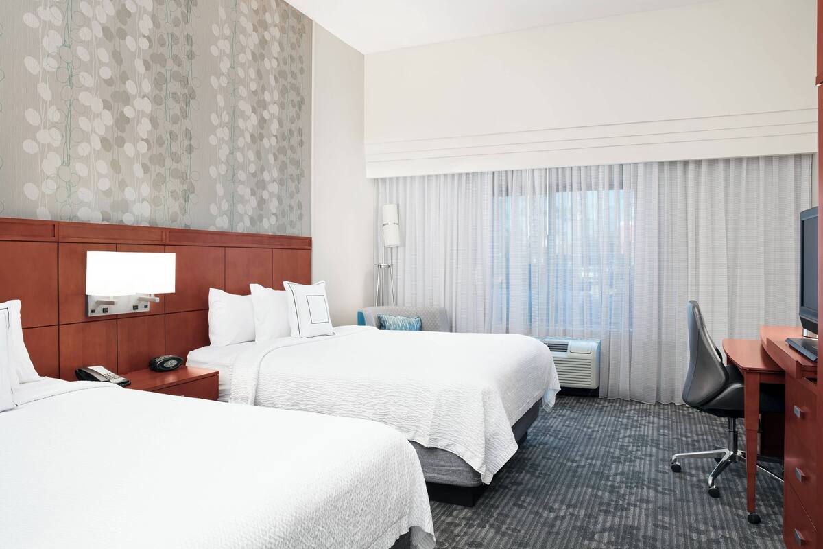 Quarto de hotel com duas camas, uma escrivaninha, uma cadeira, um abajur e uma janela com cortinas.