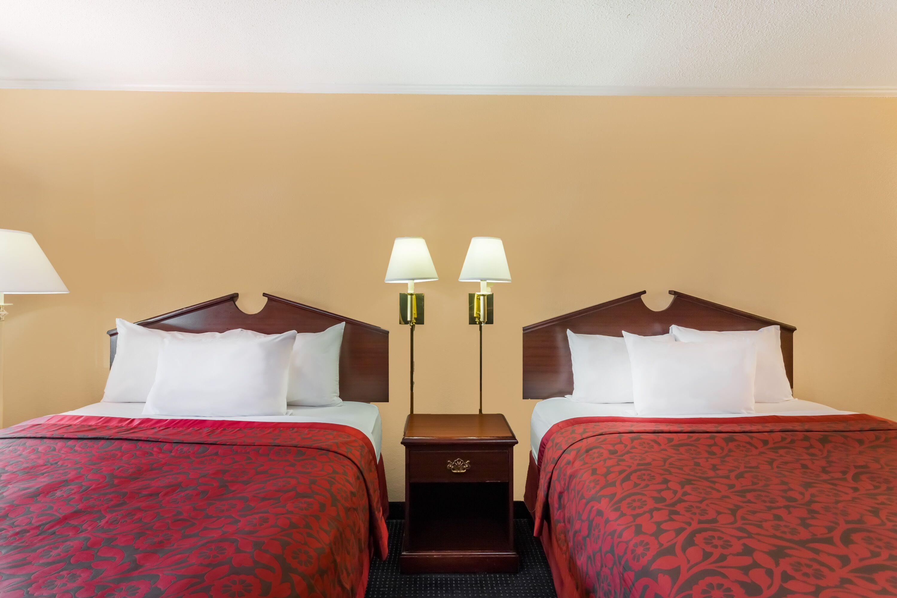 Quarto de hotel com duas camas, cada uma com cabeceira de madeira, roupa de cama branca e edredons estampados em vermelho. Há duas luminárias de cabeceira com base verde e abajures brancos.