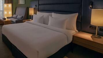 Quarto de hotel com uma cama grande, uma poltrona, duas mesas de cabeceira com luminárias e um painel de controle fixado na parede.